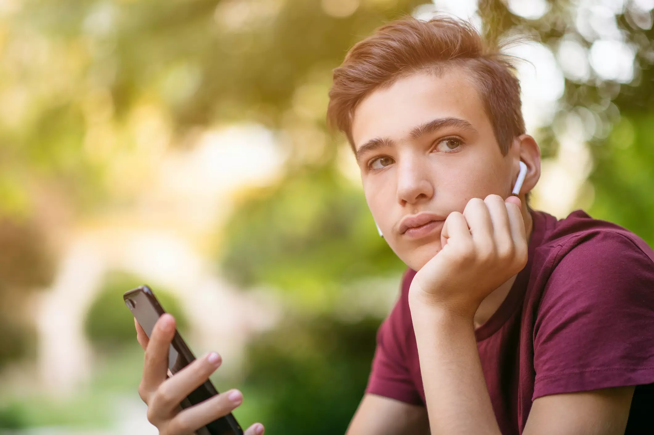 Detailný portrét zamysleného nešťastného dospievajúceho chlapca so smartfónom vonku. Smutný tínedžer s mobilným telefónom pozerá inam, v parku. Zamyslený teenager v neformálnom oblečení s mobilným telefónom v parku