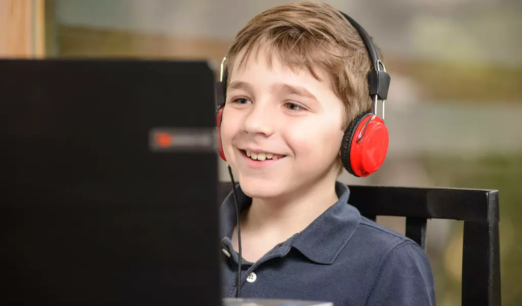 ADHD असलेला मुलगा हेडफोन घातलेला, हसत हसत आणि डेस्कवर कॉम्प्युटरवर काम करत आहे