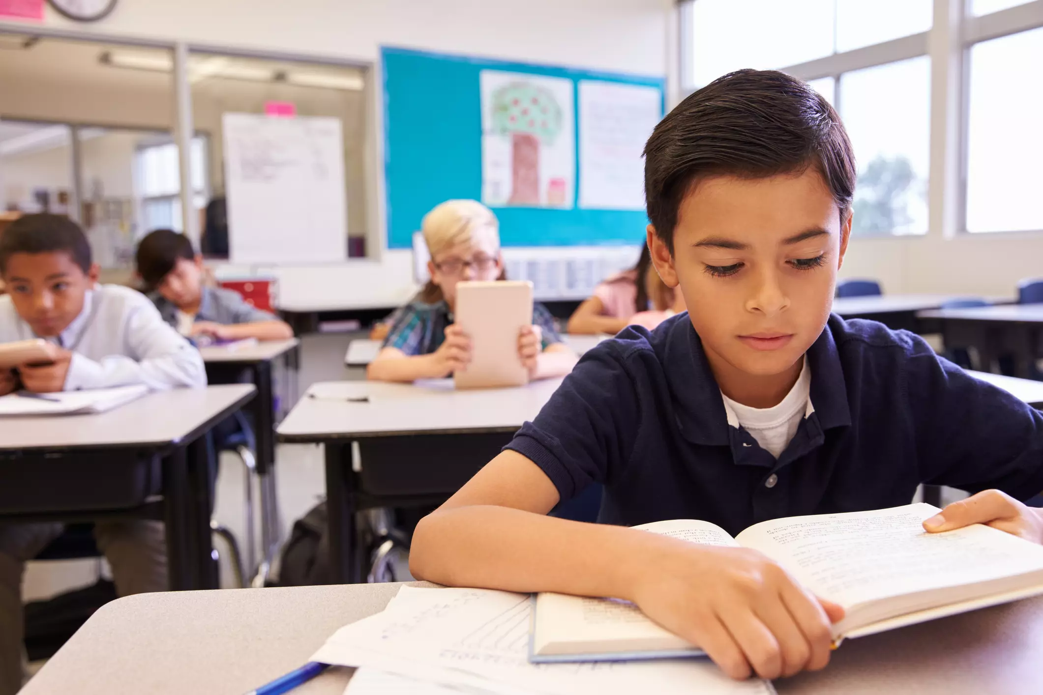 प्राथमिक शाळेच्या वर्गात ADHD शाळेचा मुलगा त्याच्या डेस्कवर वाचत आहे