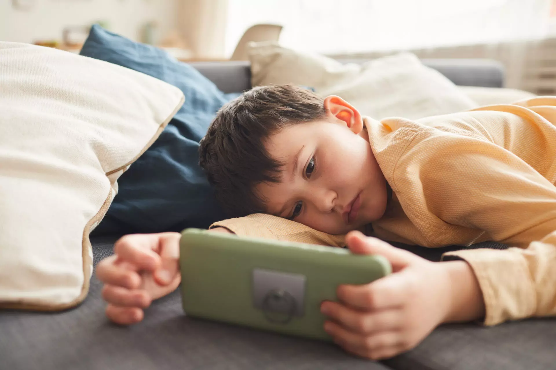 Топол портрет на досадно тинејџерско момче кое користи паметен телефон додека лежи на кревет или кауч дома, копирајте простор