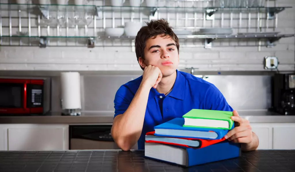 Djalë adoleshent me ADHD ulur në banakun e kuzhinës me pirg librash përpara tij duke i ngulur sytë në hapësirë