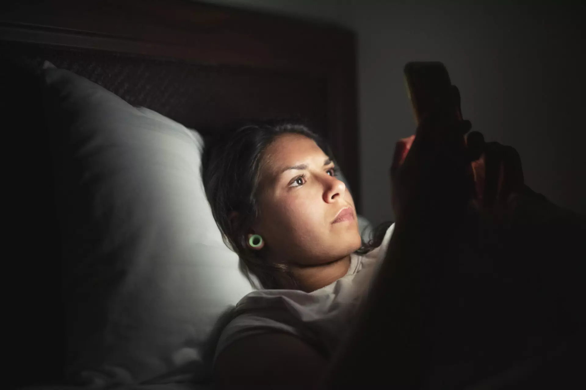 Ung kvinne som ligger i sengen og bruker smarttelefon ved lite lys.