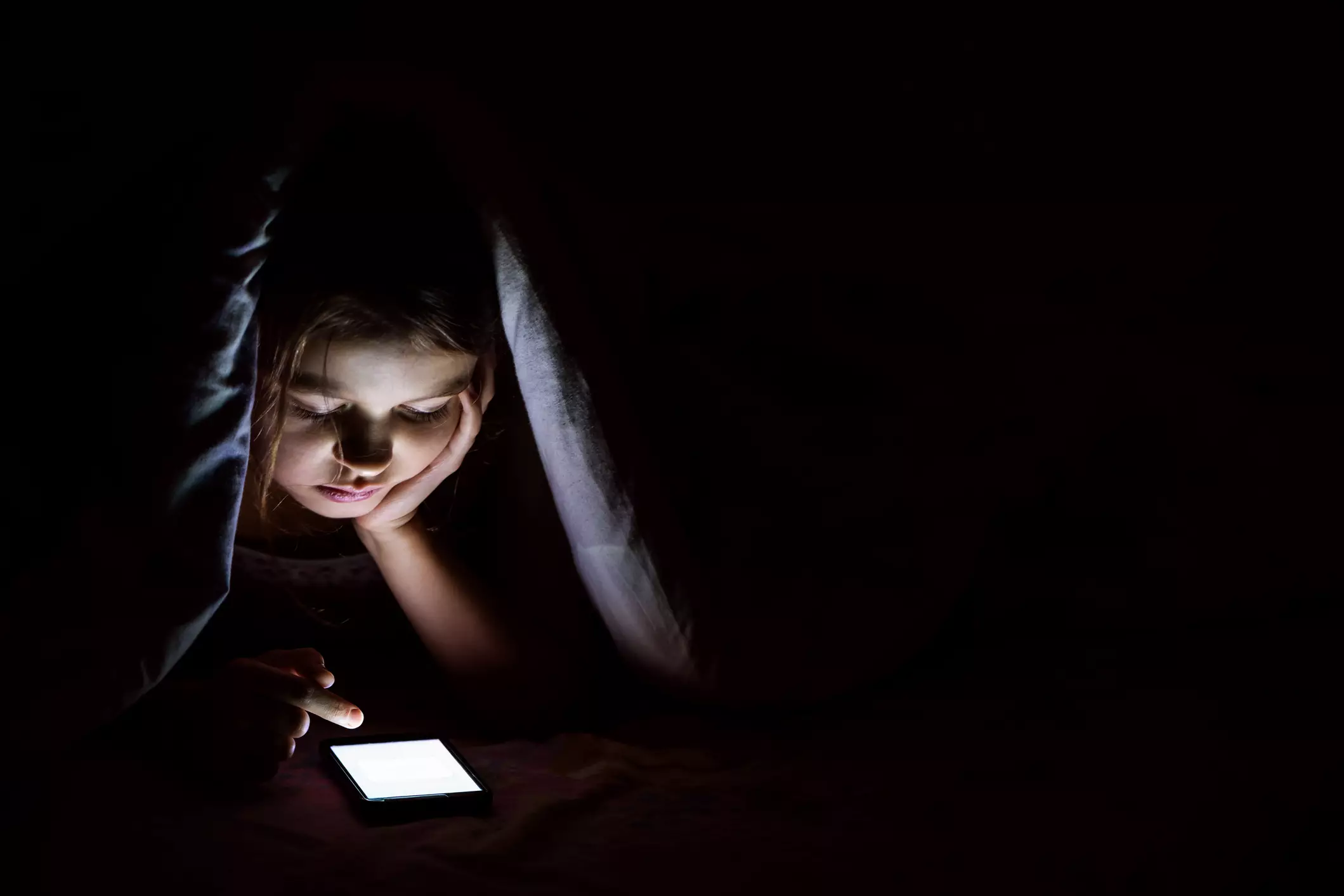 A nena de 9 anos de noite está cuberta cunha manta e mira un teléfono intelixente.