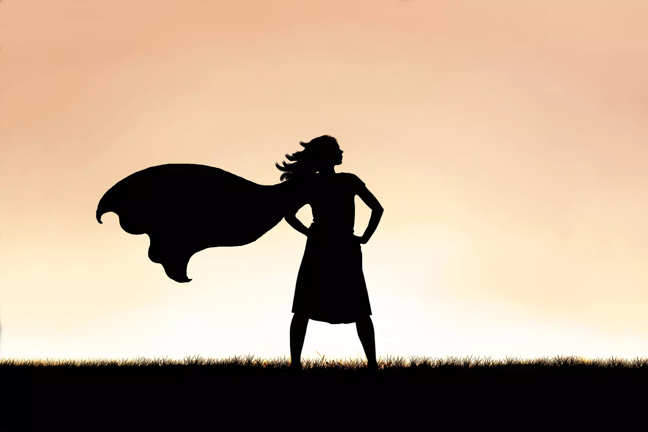 Stiprios, gražios apsiaustos super herojės moters siluetas stovi izoliuotas prieš saulėlydį dangaus fone.