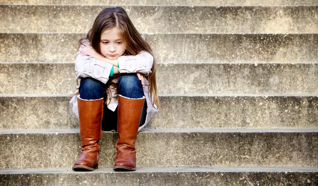 ADHD ધરાવતી છોકરી ઘૂંટણ પર હાથ ફોલ્ડ કરીને બહાર પથ્થરના પગથિયાં પર બેઠી છે