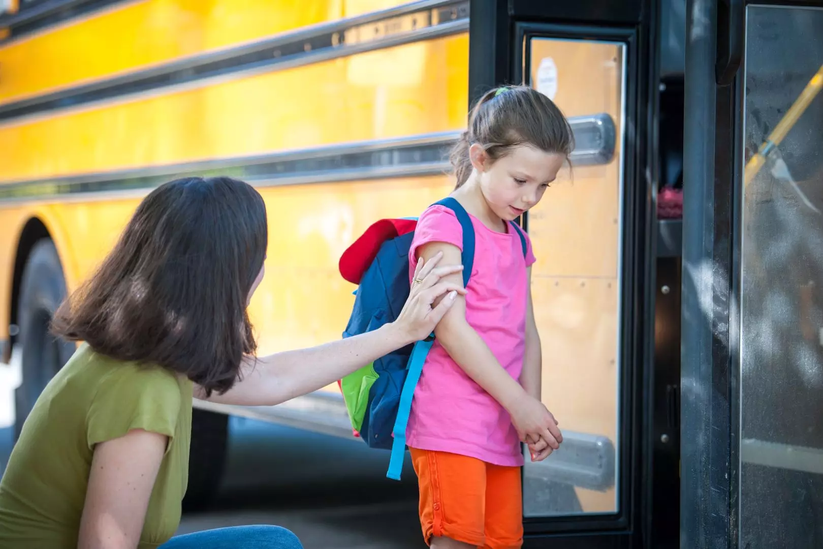 Vaikų, sergančių ADHD, nerimas: jauna mergina sulenkusi pečius atrodo drovi įlipti į mokyklinį autobusą, o mama ją guodžia liesdama ranką