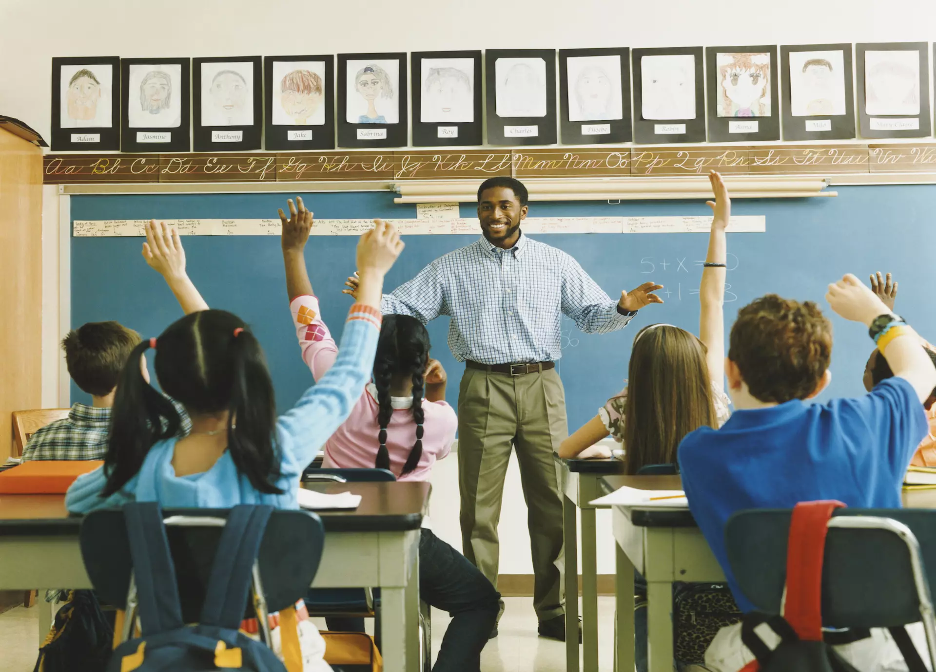 हात उंचावलेल्या वर्गासमोर उभा असलेला शिक्षक