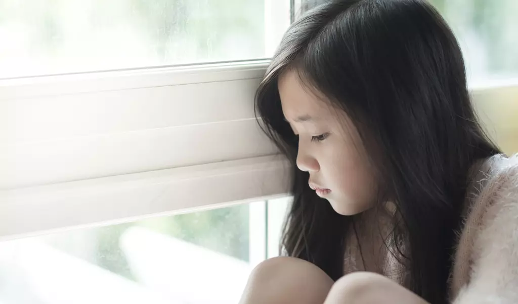 ADHD आणि नैराश्य असलेली मुलगी घराच्या खिडकीजवळ बसून भुसभुशीत आहे