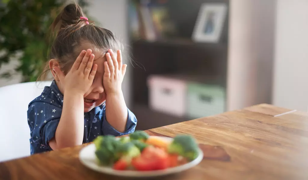 Menina com TDAH cobre o rosto com a mão enquanto está sentado na frente do prato de legumes