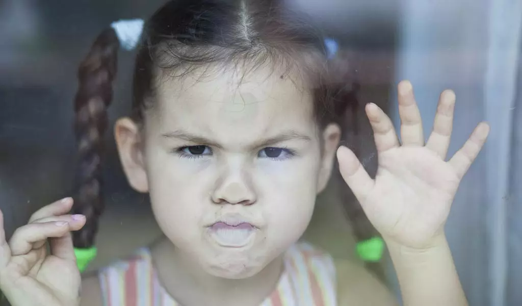 ADHD والی چھوٹی لڑکی غصے سے شیشے کے دروازے سے چہرہ دبا رہی ہے۔