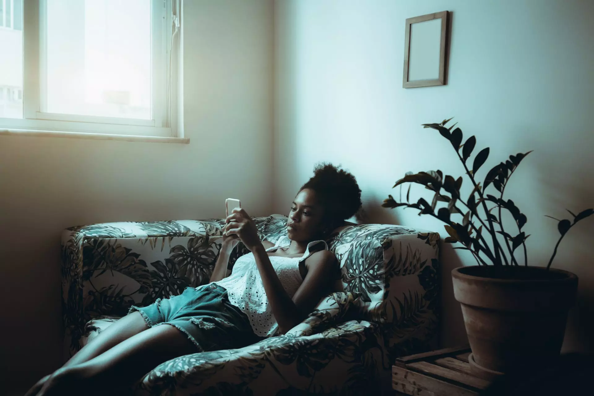 امرأة شابة من أصل أفريقي لطيفة تتصفح صفحات الإنترنت على هاتف ذكي بينما تستلقي على الأريكة بالقرب من النافذة والنبات ، وتشعر بالملل من الحجر الصحي والعزلة