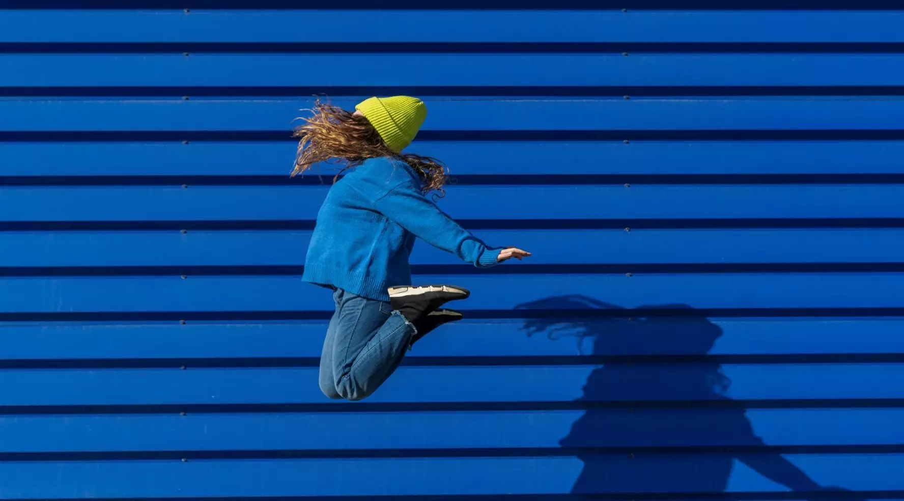 Sınırlayıcı inançlarınızdan kurtulun: Sarı bir bere takan genç bir kız mavi bir zemine karşı yükseğe zıplıyor.